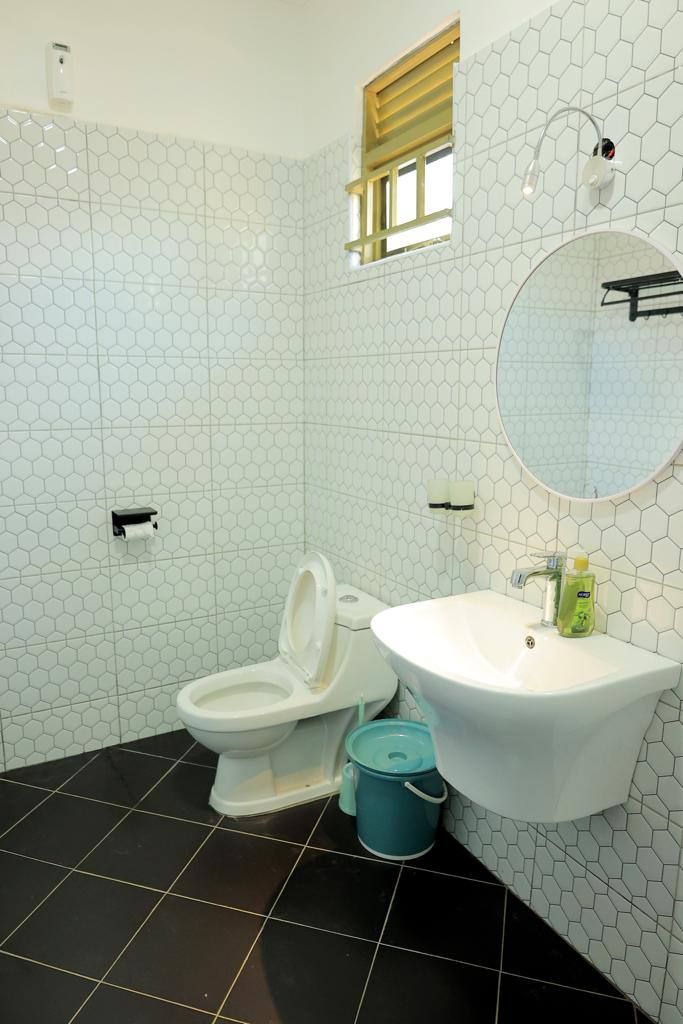 Casa de banho com sanita, lavatório e espelho. Há uma janela sobre a sanita, e um balde azul está no chão.