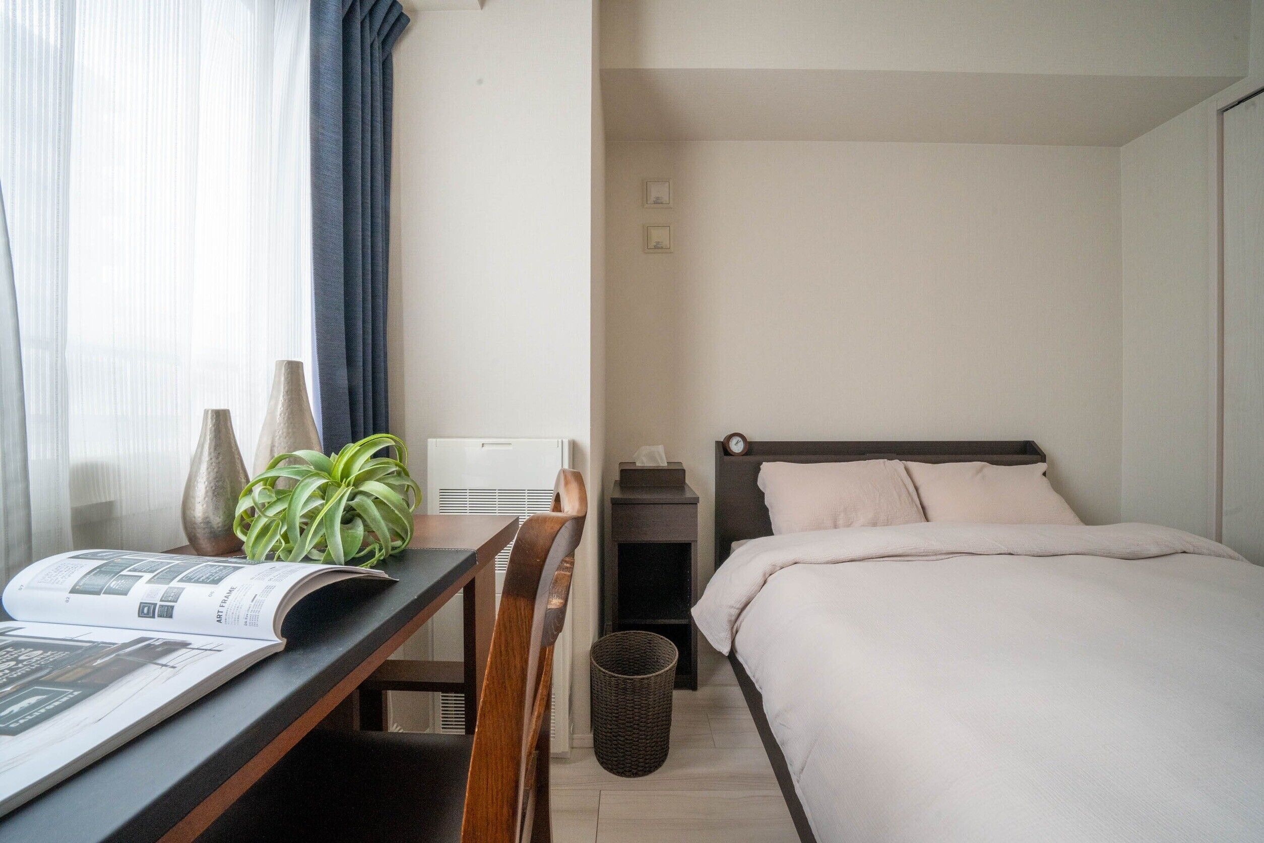 Quarto de hotel bem arrumado com cama, mesa com uma revista, planta em vaso e lixeira.