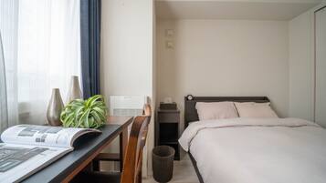 Quarto de hotel bem arrumado com cama, mesa com uma revista, planta em vaso e lixeira.