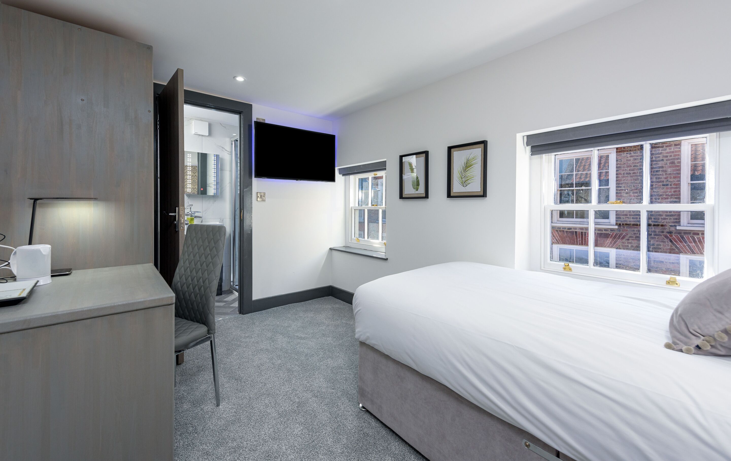 Quarto moderno com cama, escrivaninha, cadeira, televisão e janela com vista para um edifício de tijolos.