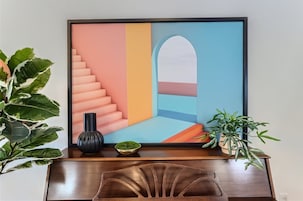 Pintura abstrata emoldurada, com formas geométricas e design de escada, apoiada em uma mesa de madeira com um vaso e uma pequena planta.