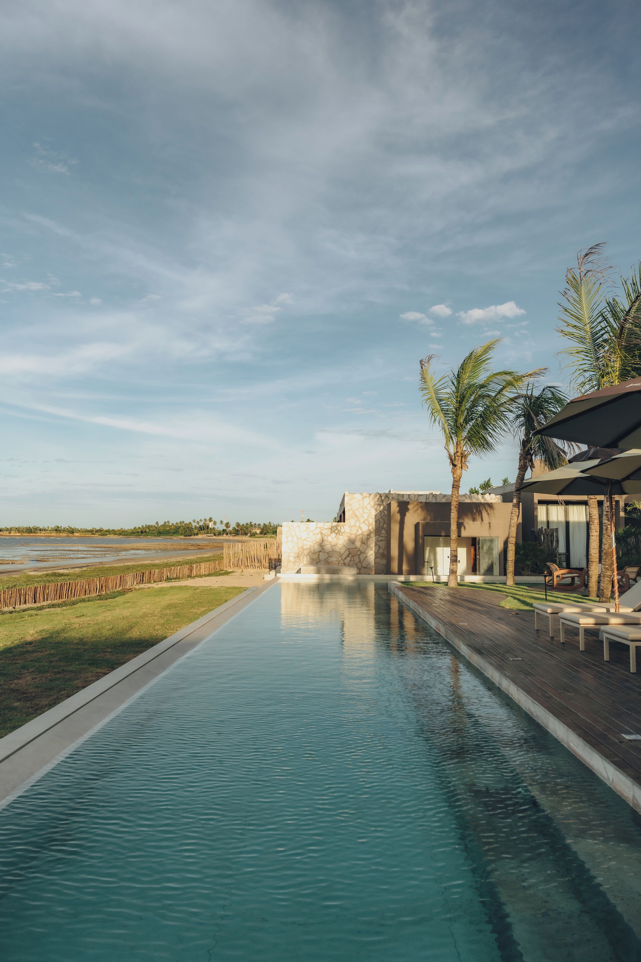 Uma área de piscina com vista para o oceano, palmeiras e um edifício com fachada de pedra.