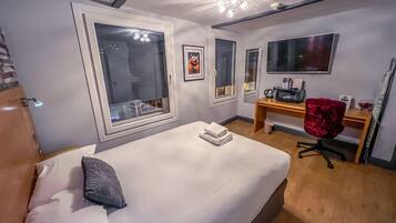 Quarto de hotel com cama, escrivaninha, cadeira, televisão e janela.