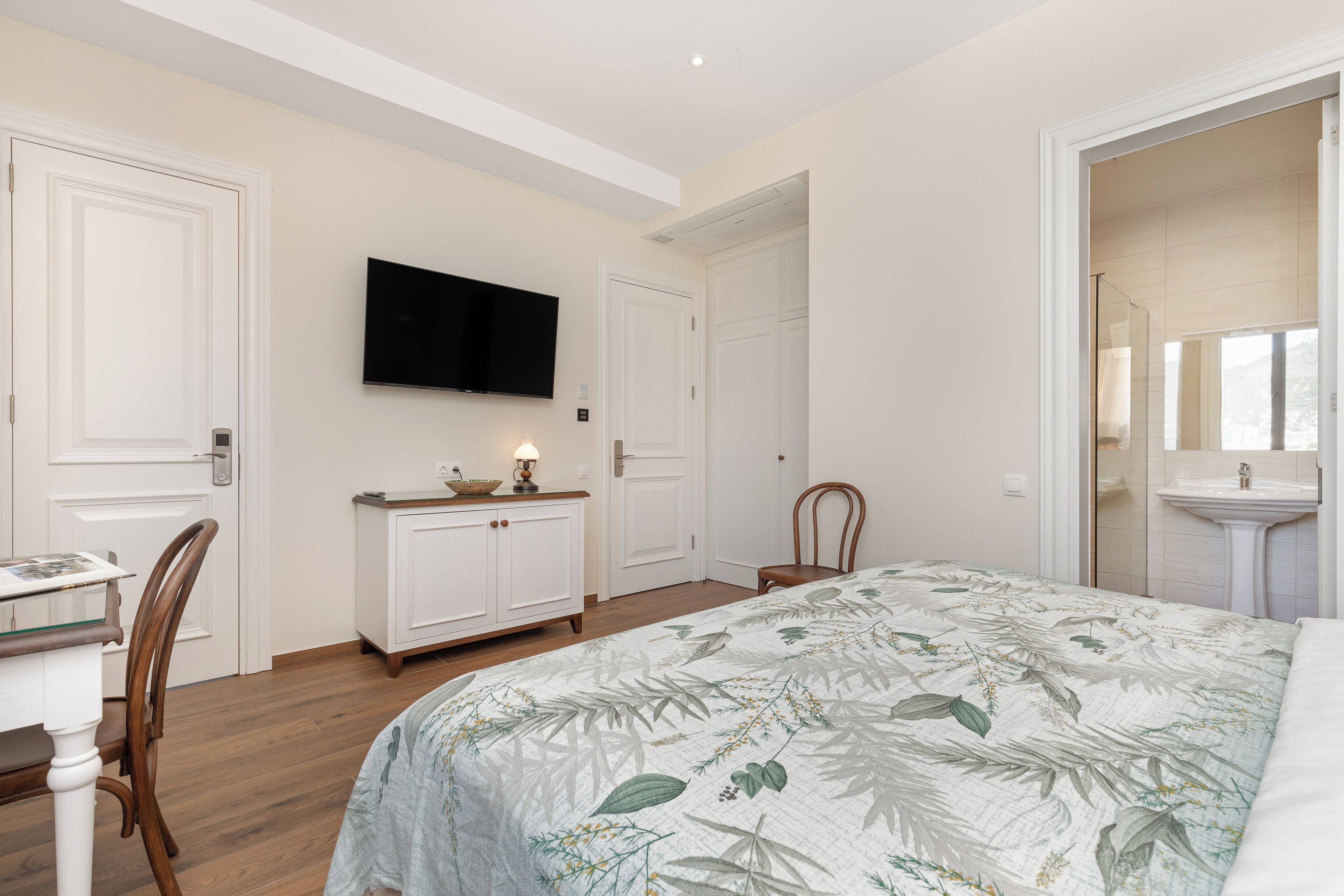 Quarto com cama, televisão fixada na parede, escrivaninha com cadeira e banheiro com pia e espelho.