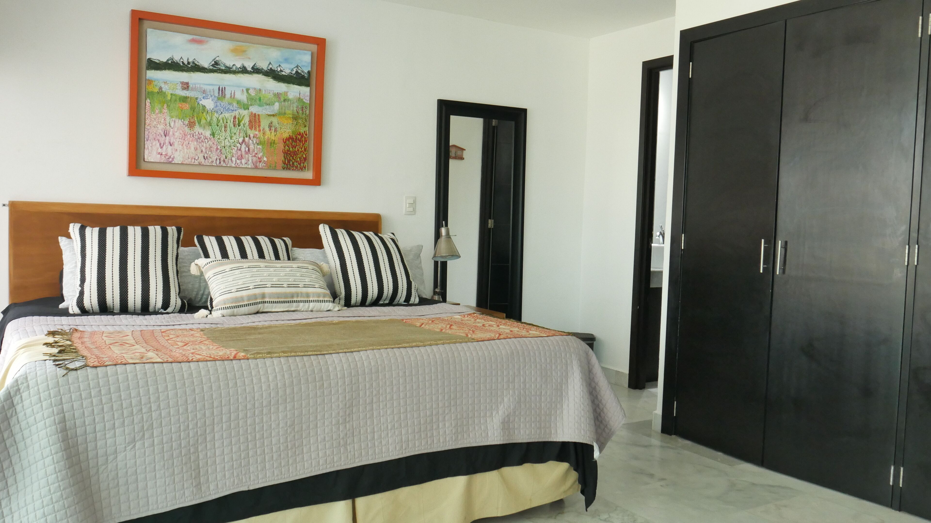Quarto com cama, almofadas, abajur, quadro e guarda-roupa.
