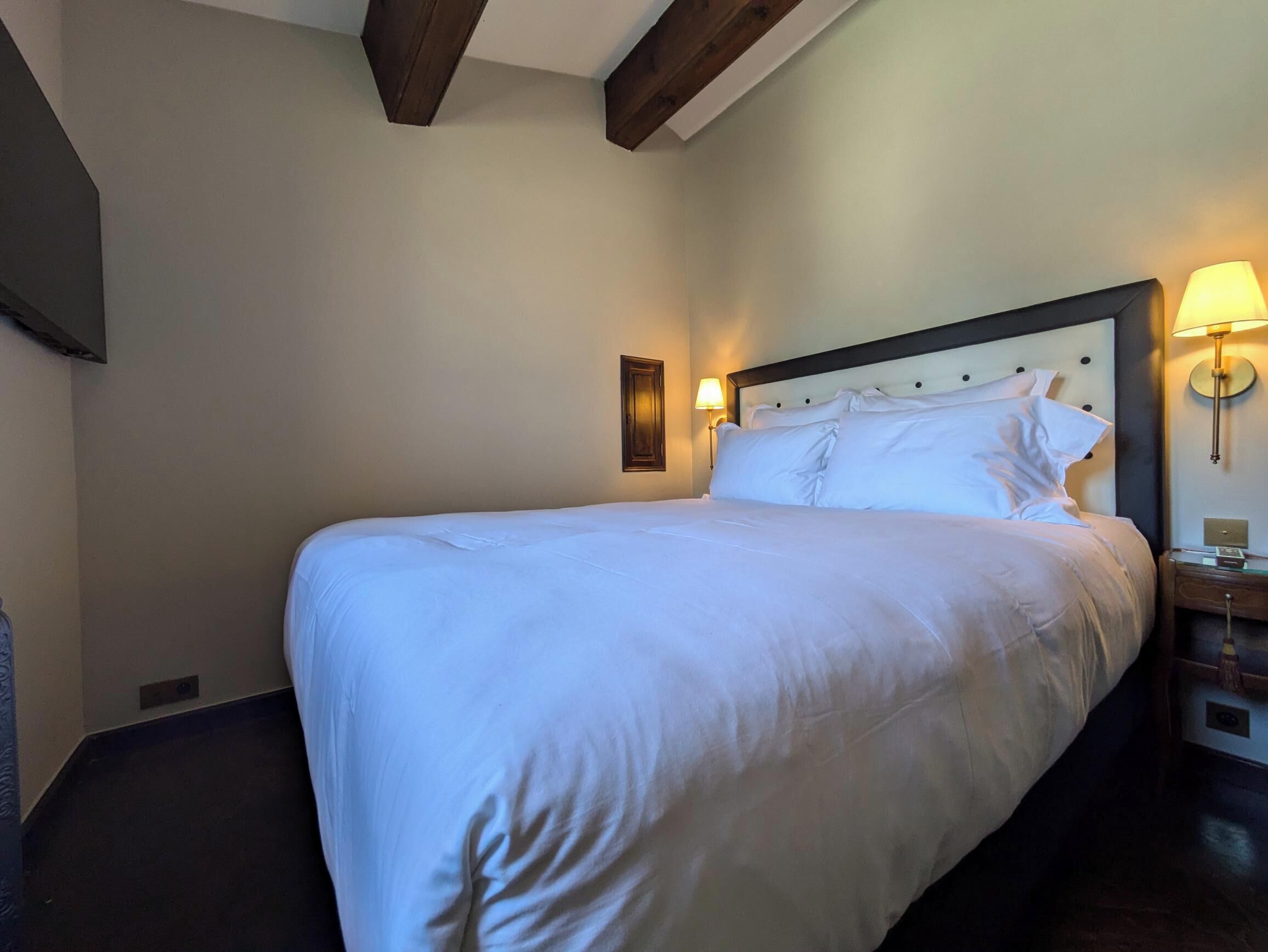 Uma cama bem arrumada em quarto de hotel, com cabeceira de madeira, abajur na mesinha de cabeceira e uma pequena mesa com luminária ao lado.