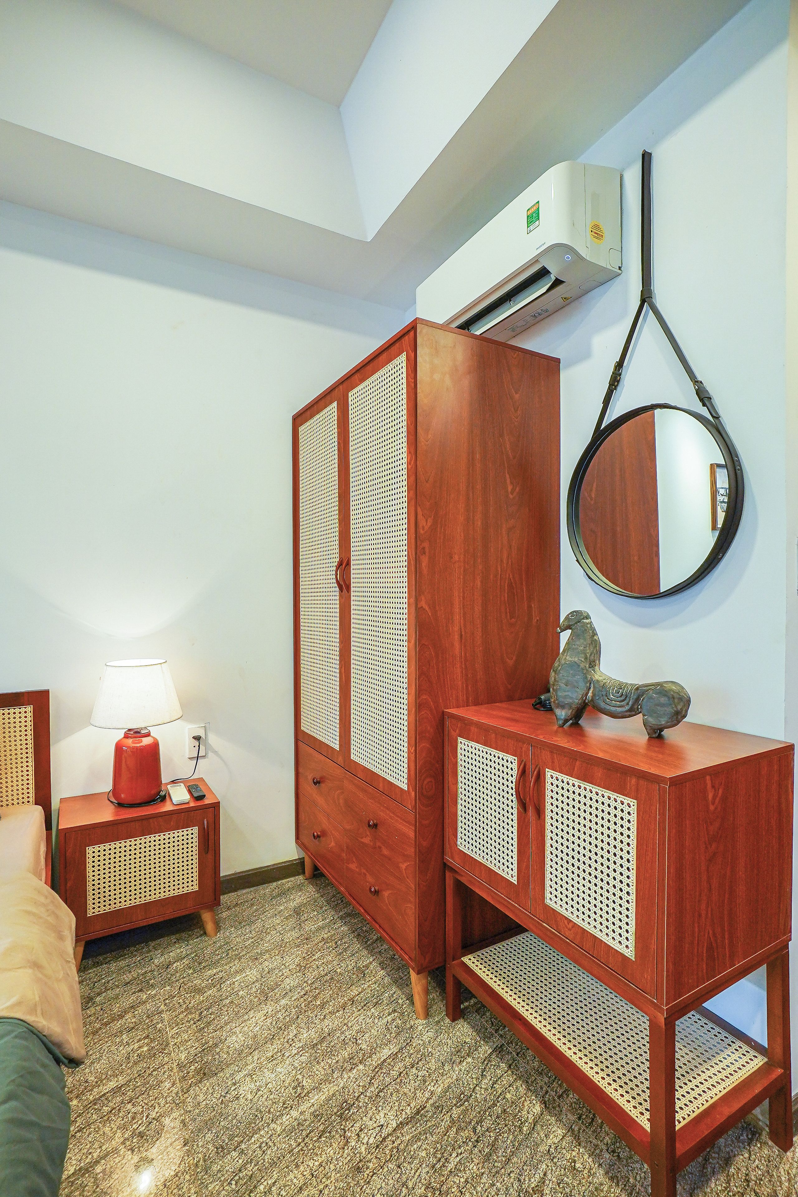 Quarto de hotel com guarda-roupa de madeira, espelho redondo, pequena mesa com abajur e chão coberto por carpete.