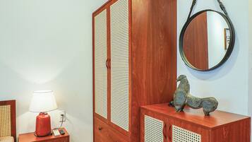 Quarto de hotel com guarda-roupa de madeira, espelho redondo, pequena mesa com abajur e chão coberto por carpete.