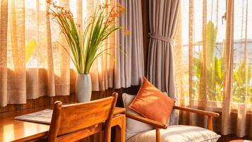 Quarto com mesa e cadeiras de madeira, planta em vaso e vista através de janelas amplas com cortinas.