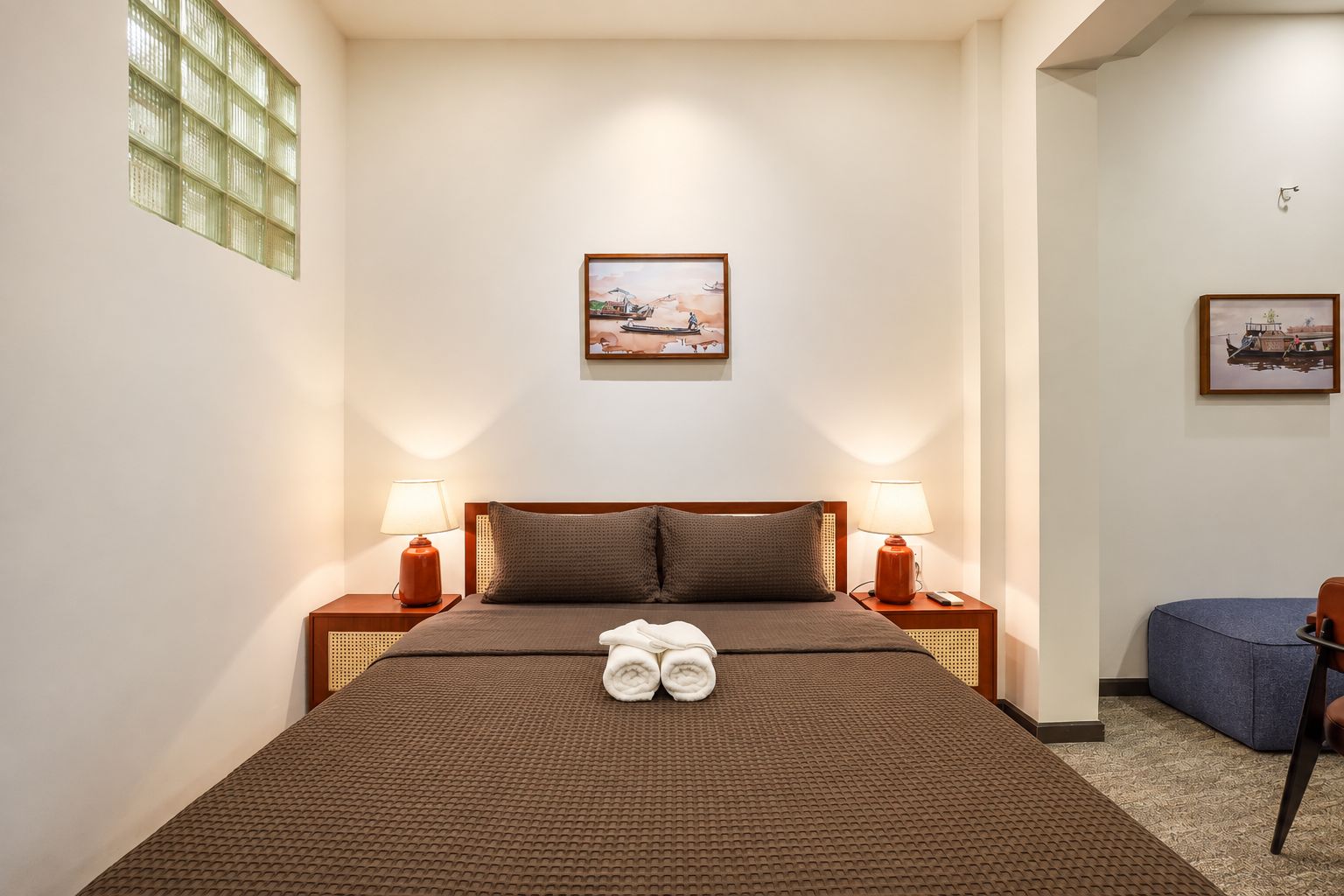 Quarto de hotel com uma cama grande, duas luminárias de cabeceira, um quadro na parede e uma pequena área de estar com uma poltrona.