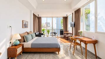 Quarto de hotel com cama, escrivaninha, cadeiras, televisão e grandes janelas.