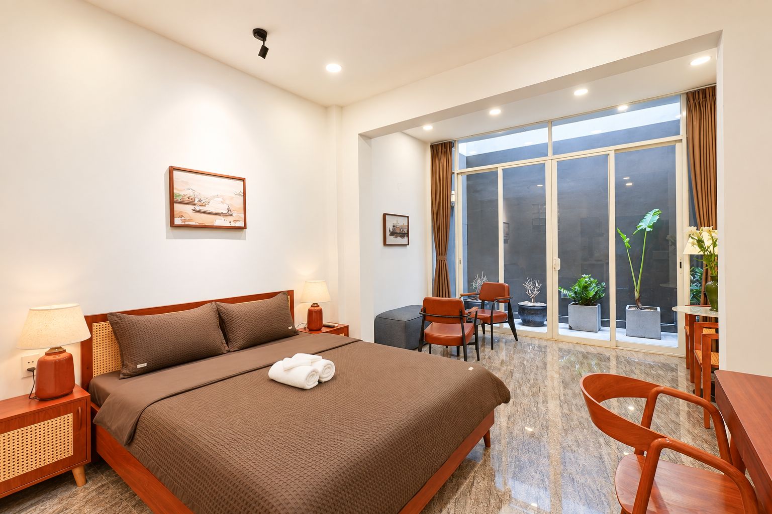 Quarto de hotel com cama grande, duas mesas de cabeceira com abajures, área de estar com cadeiras e mesa, e vista pela grande janela.