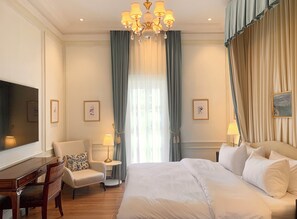 Quarto de hotel com uma cama grande, uma escrivaninha, uma cadeira, uma mesinha de cabeceira com uma luminária e uma televisão na parede.