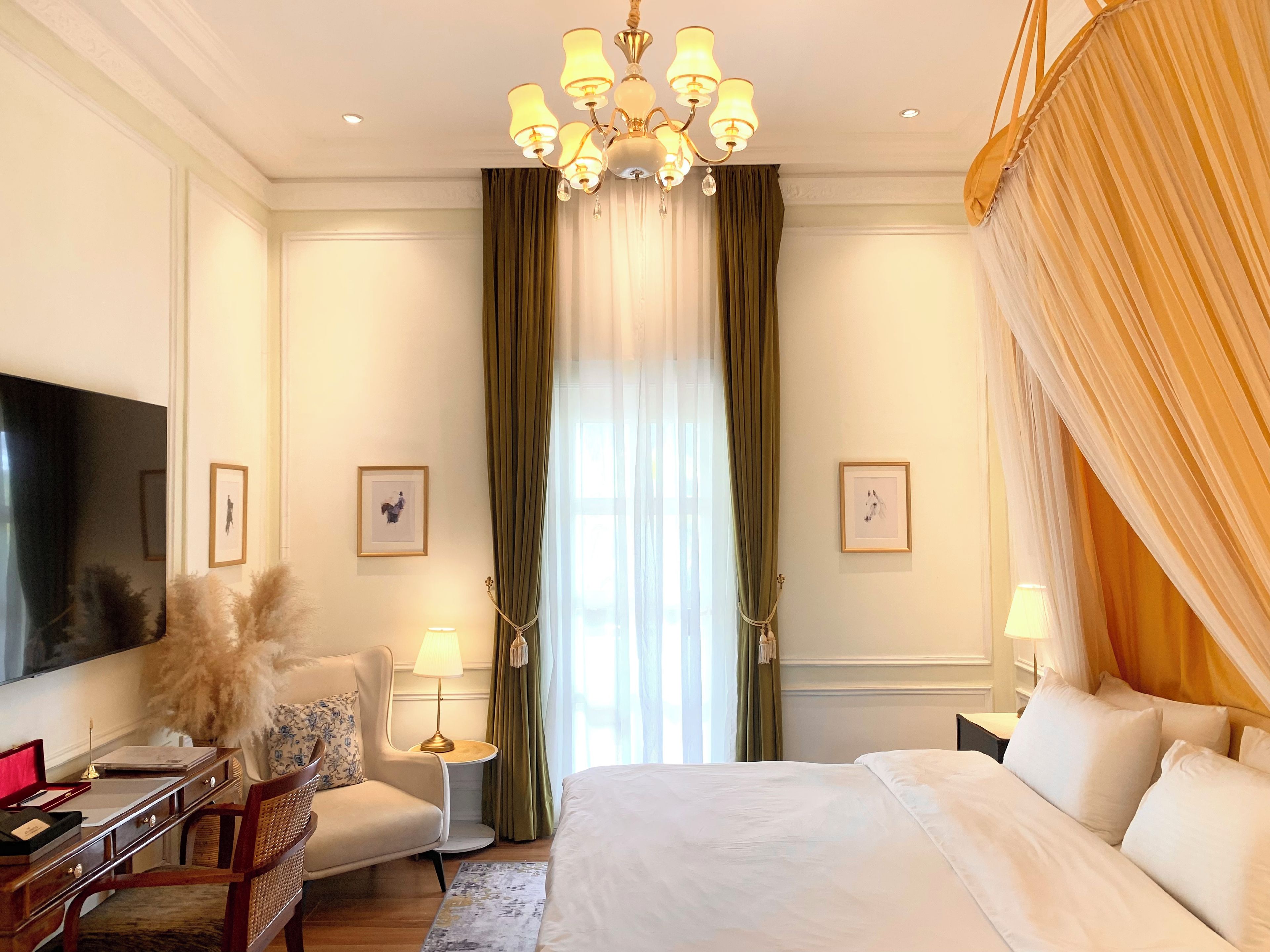 Quarto de hotel com uma cama grande, uma escrivaninha com luminária, uma cadeira, uma televisão e um lustre.