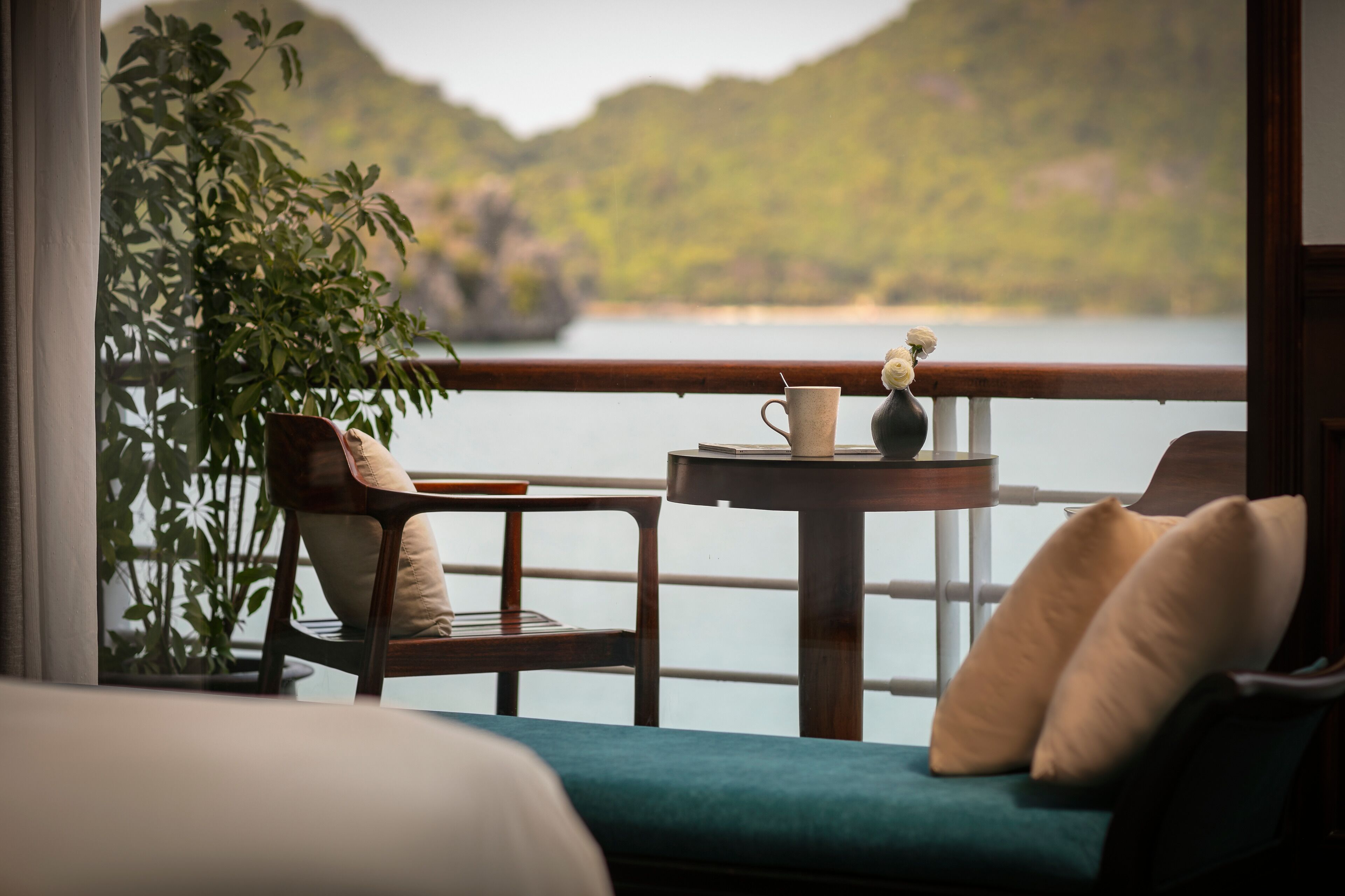 Uma varanda com vista para um lago, uma mesa com uma xícara e um vaso, e uma cadeira com almofadas.