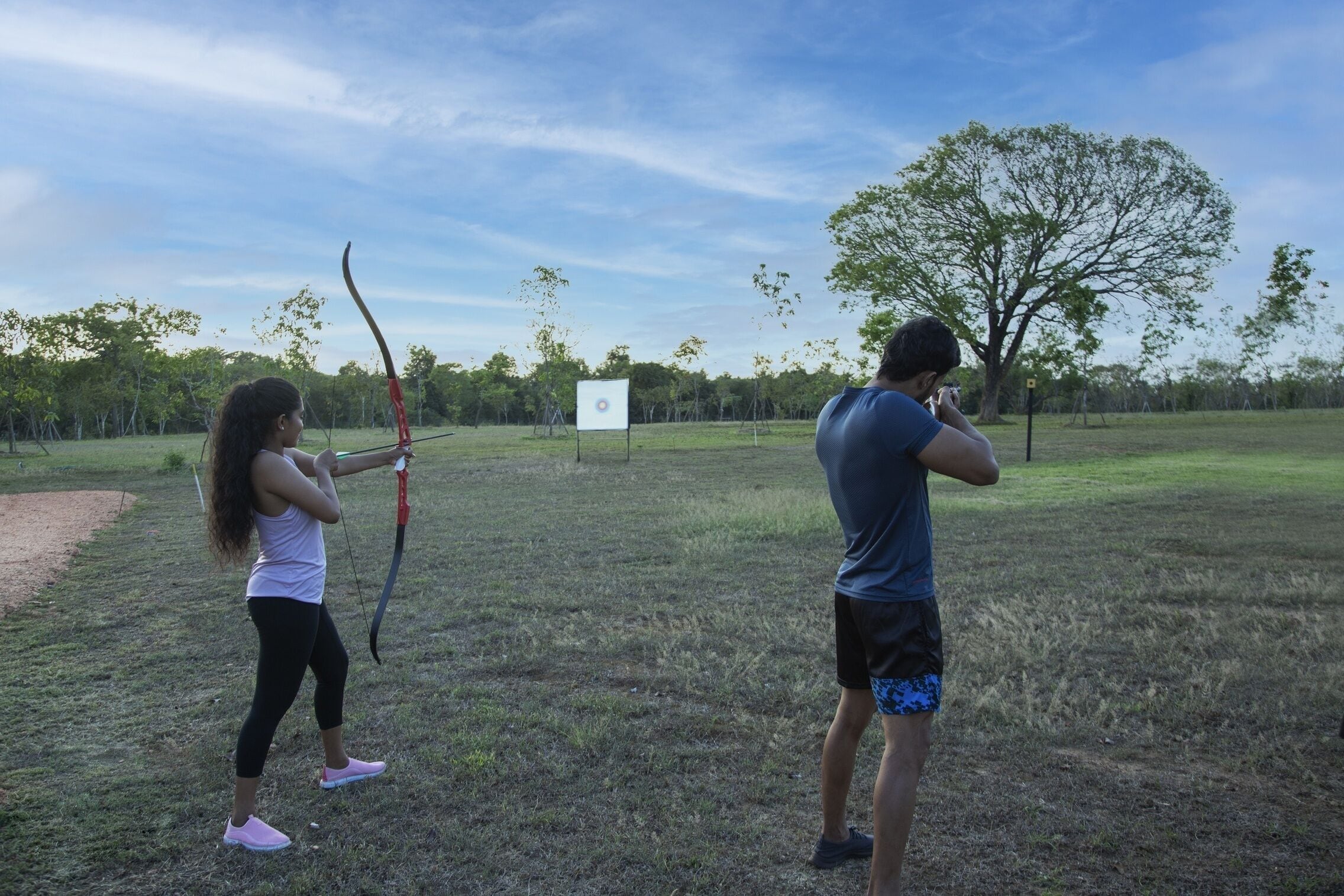 Duas pessoas praticando arco e flecha em um campo aberto, com um alvo ao longe.