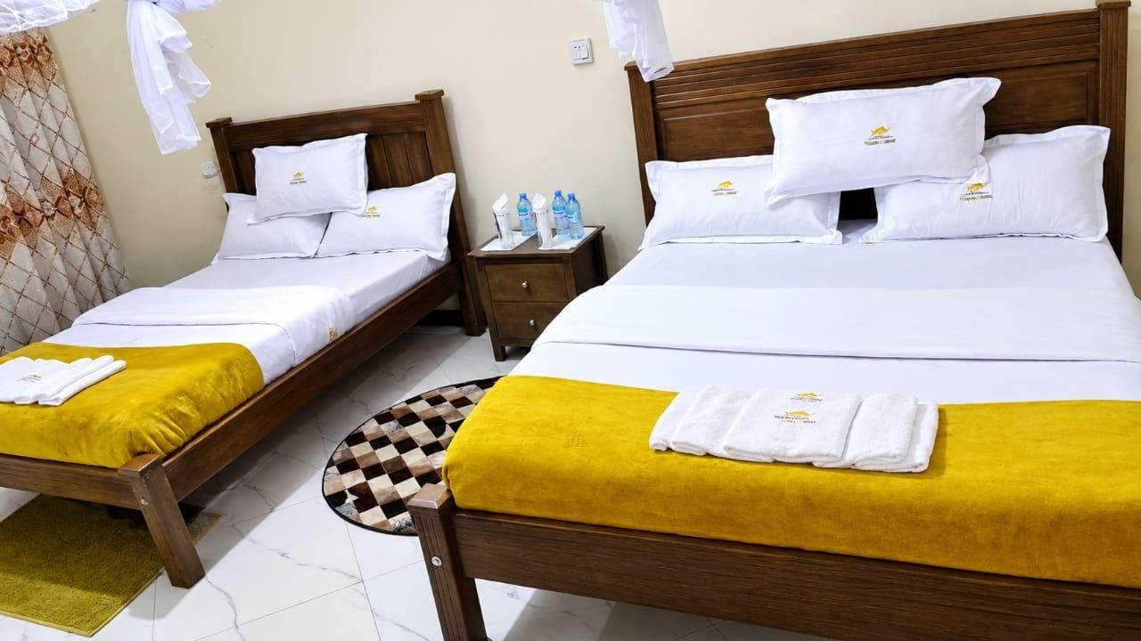 Quarto de hotel com duas camas, cada uma com roupa de cama branca e almofadas a combinar. Há uma pequena mesa de cabeceira com uma garrafa de água e uma toalha.