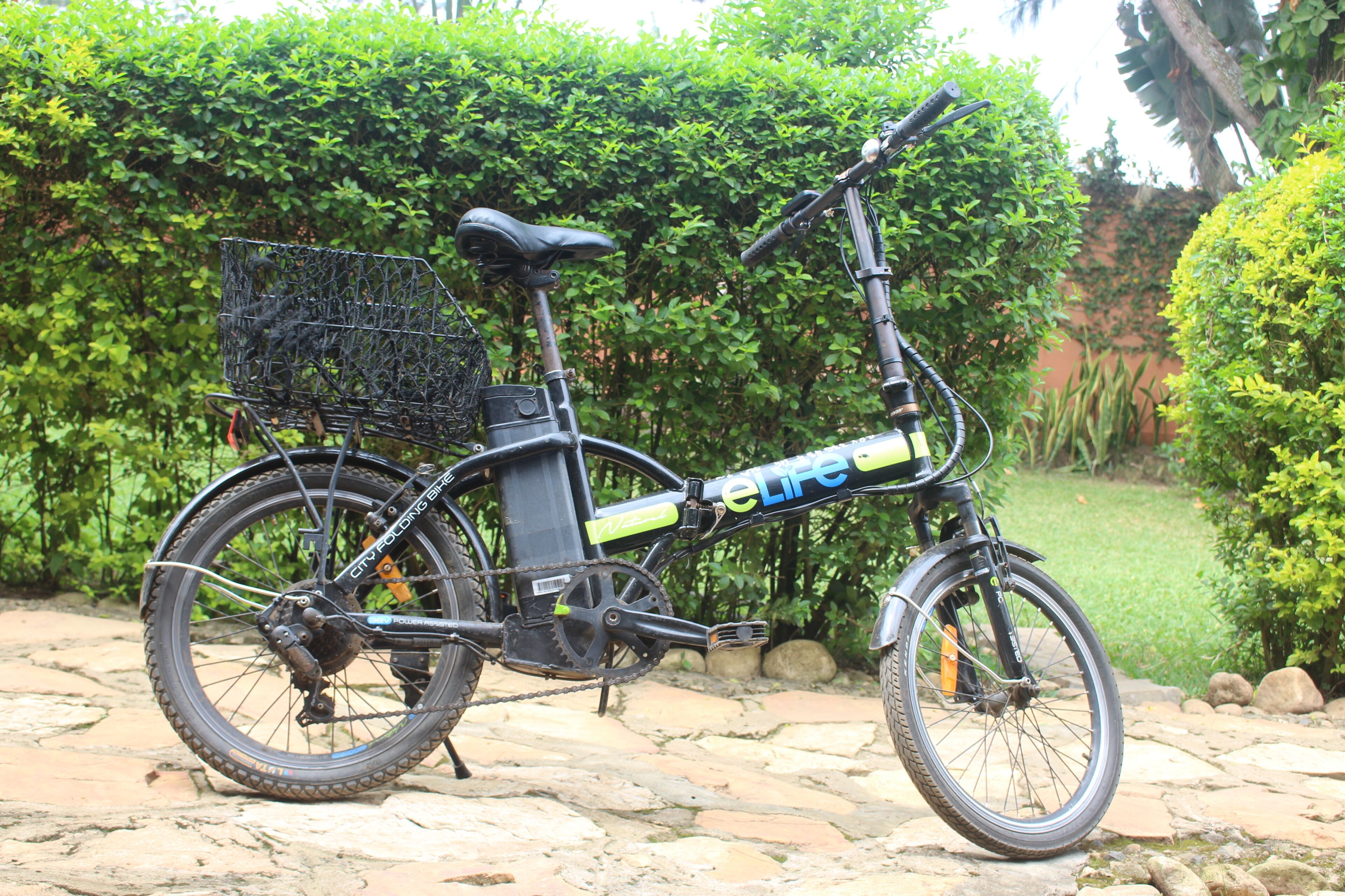 Uma bicicleta ergométrica com cesto e garrafa de água, estacionada em um caminho de pedra.