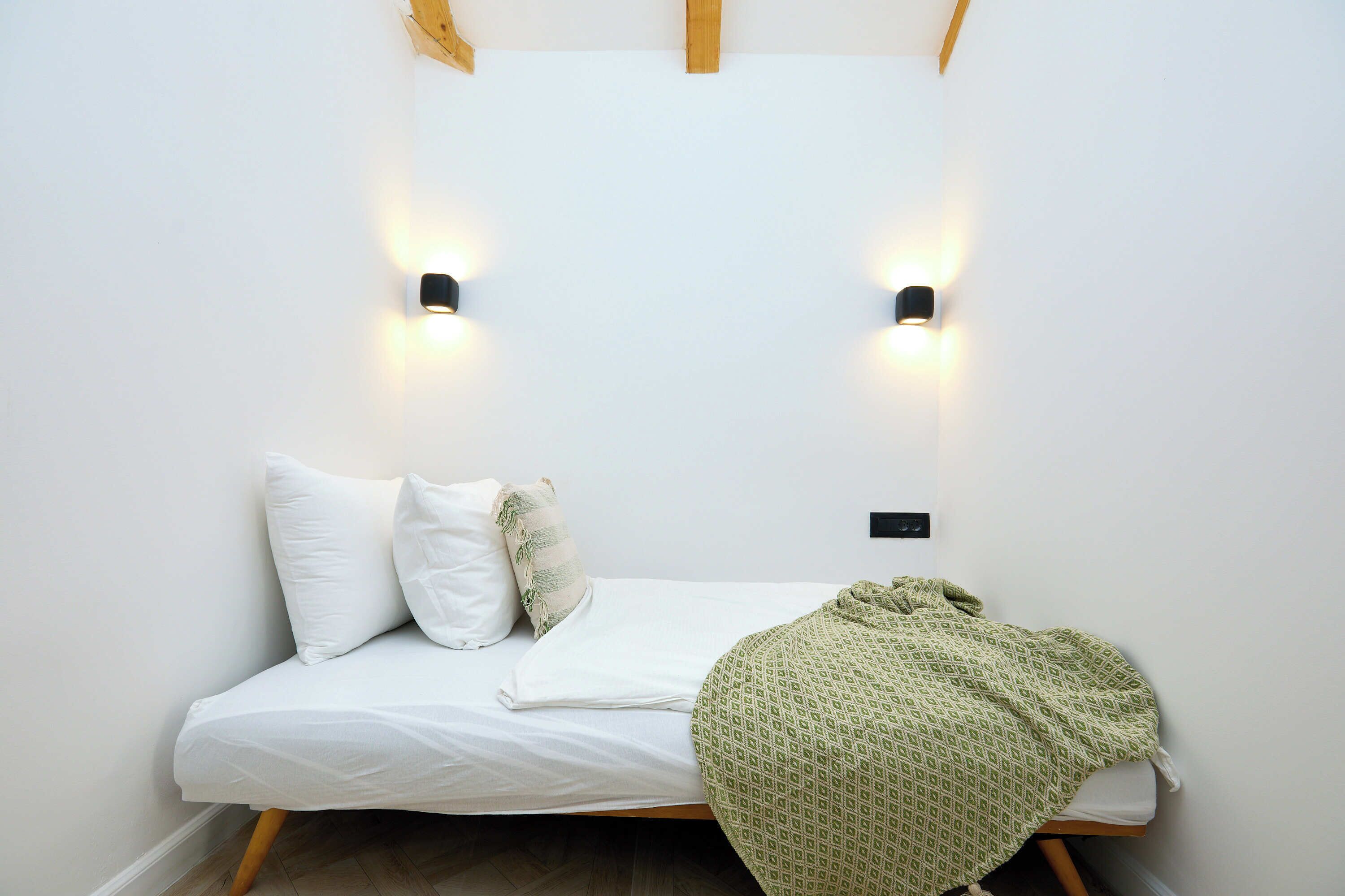 Uma cama pequena e bem arrumada com roupa de cama branca e um cobertor verde estampado, duas luminárias fixadas na parede e uma cabeceira de madeira.