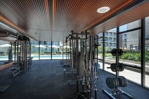 Academia com equipamento de musculação, bancos e vista para uma piscina e edifícios.