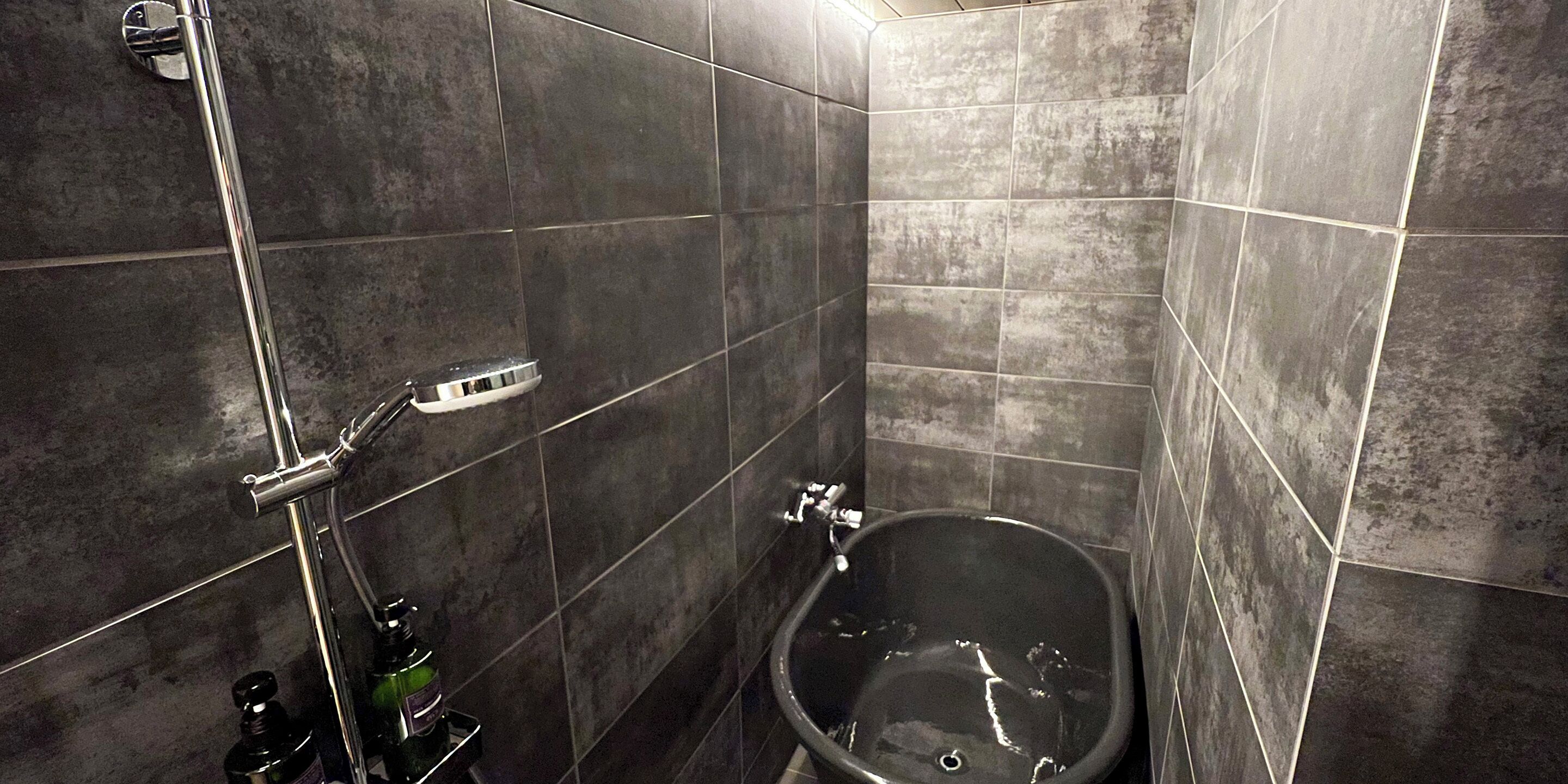 Casa de banho com azulejos escuros, uma banheira e um chuveiro com torneira cromada.