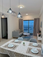 Sala de estar moderna com área de jantar, sofá cinza e vista para o horizonte urbano através de amplas janelas.