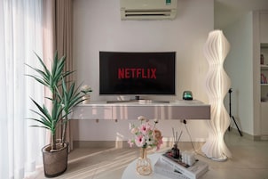 Uma sala de estar moderna com uma televisão de tela plana exibindo "NETFLIX", uma planta em vaso, um vaso decorativo e um abajur branco exclusivo.