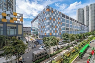 Um edifício moderno com fachada geométrica, rodeado por vegetação e uma rua movimentada.