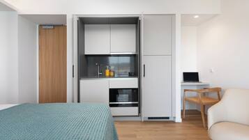 Quarto compacto com cama, cozinha compacta, uma cadeira e mesa com um laptop.