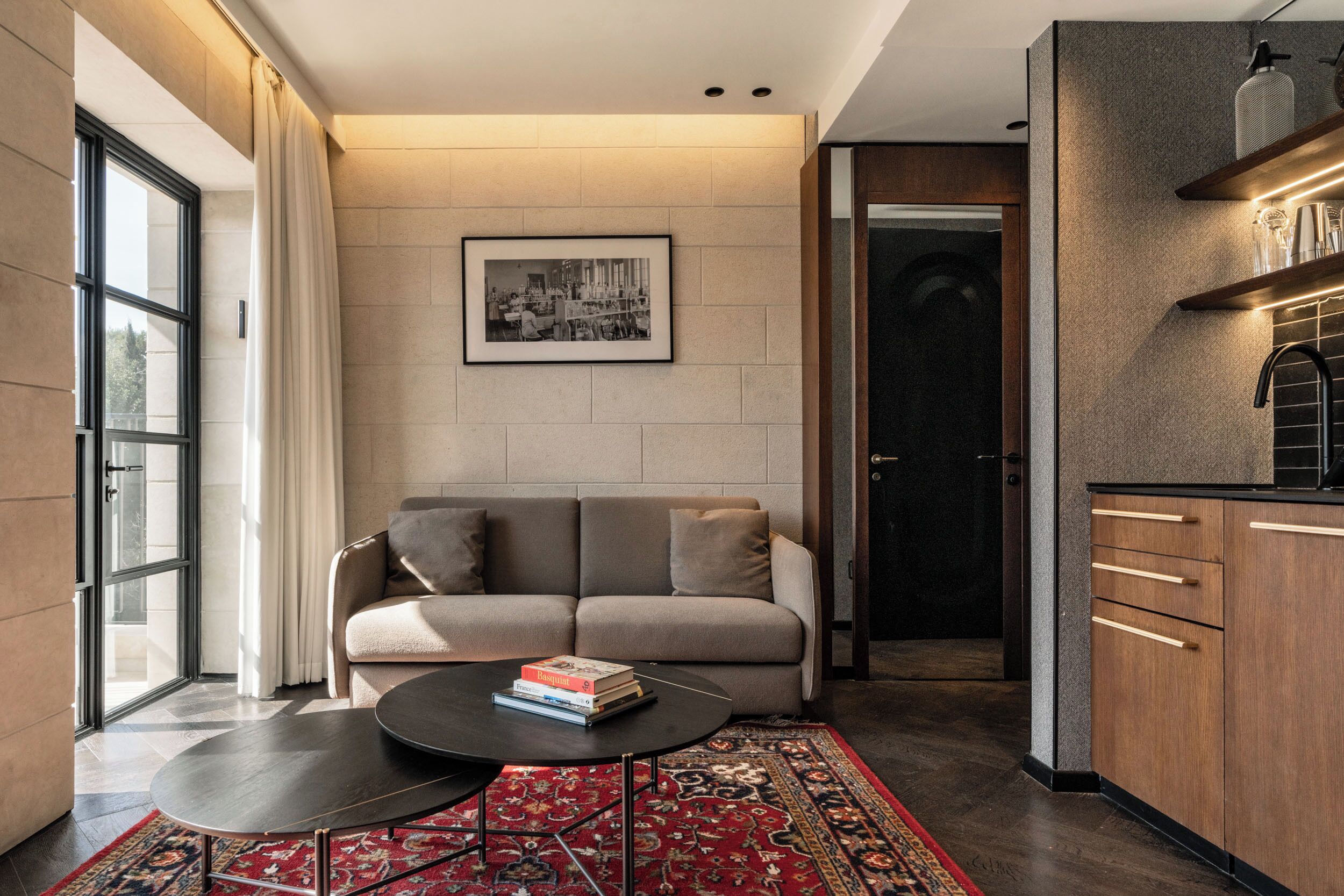 Uma sala de estar moderna com um sofá, uma mesa de centro e um tapete. Há um quadro na parede e uma área de cozinha com armários de madeira.