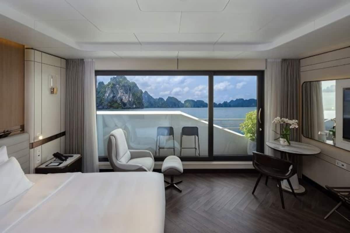 Quarto de hotel com cama, escrivaninha, cadeira, mesa pequena com um vaso e uma janela grande com vista para o mar.