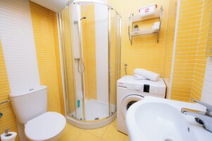 Casa de banho com chuveiro em cabine de vidro, sanita, máquina de lavar roupa e lavatório.