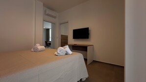 Quarto de hotel com cama, escrivaninha, televisão e banheiro visível através de uma porta aberta.