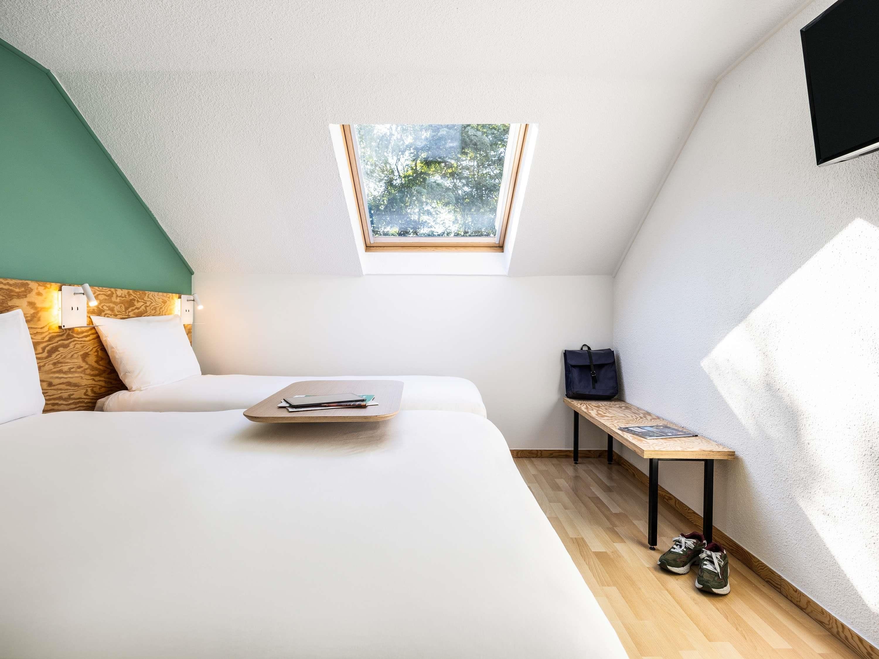 Um quarto moderno com teto inclinado, claraboia, cama com roupa de cama branca, mesa de cabeceira com um livro e uma bolsa, e um par de sapatos no chão.