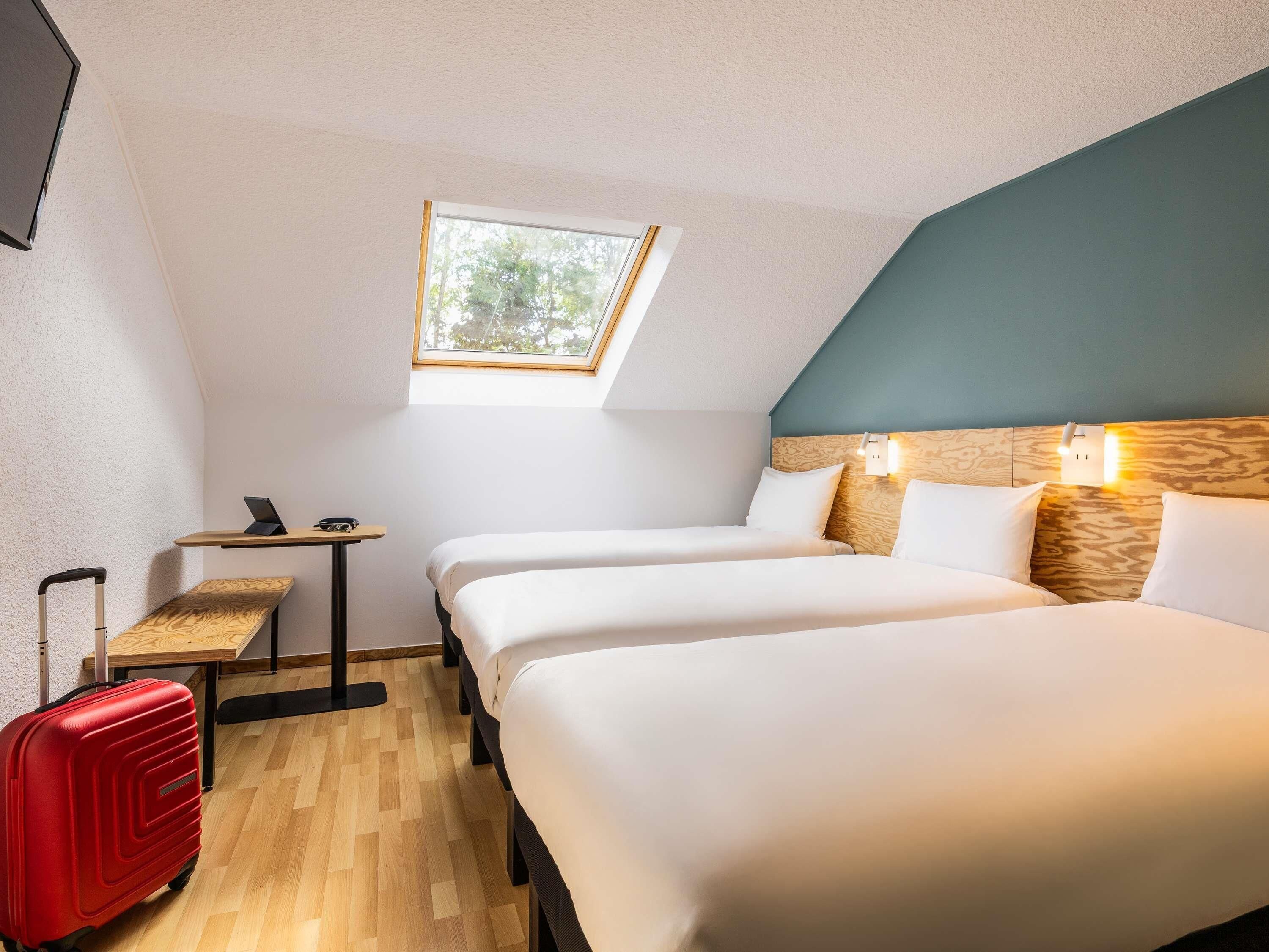 Quarto de hotel pequeno com duas camas, uma mesa, escrivaninha, janela e mala vermelha.