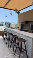 Uma área de bar ao ar livre com balcão de concreto, banquetas de madeira, luminárias pendentes e vista para a cidade.