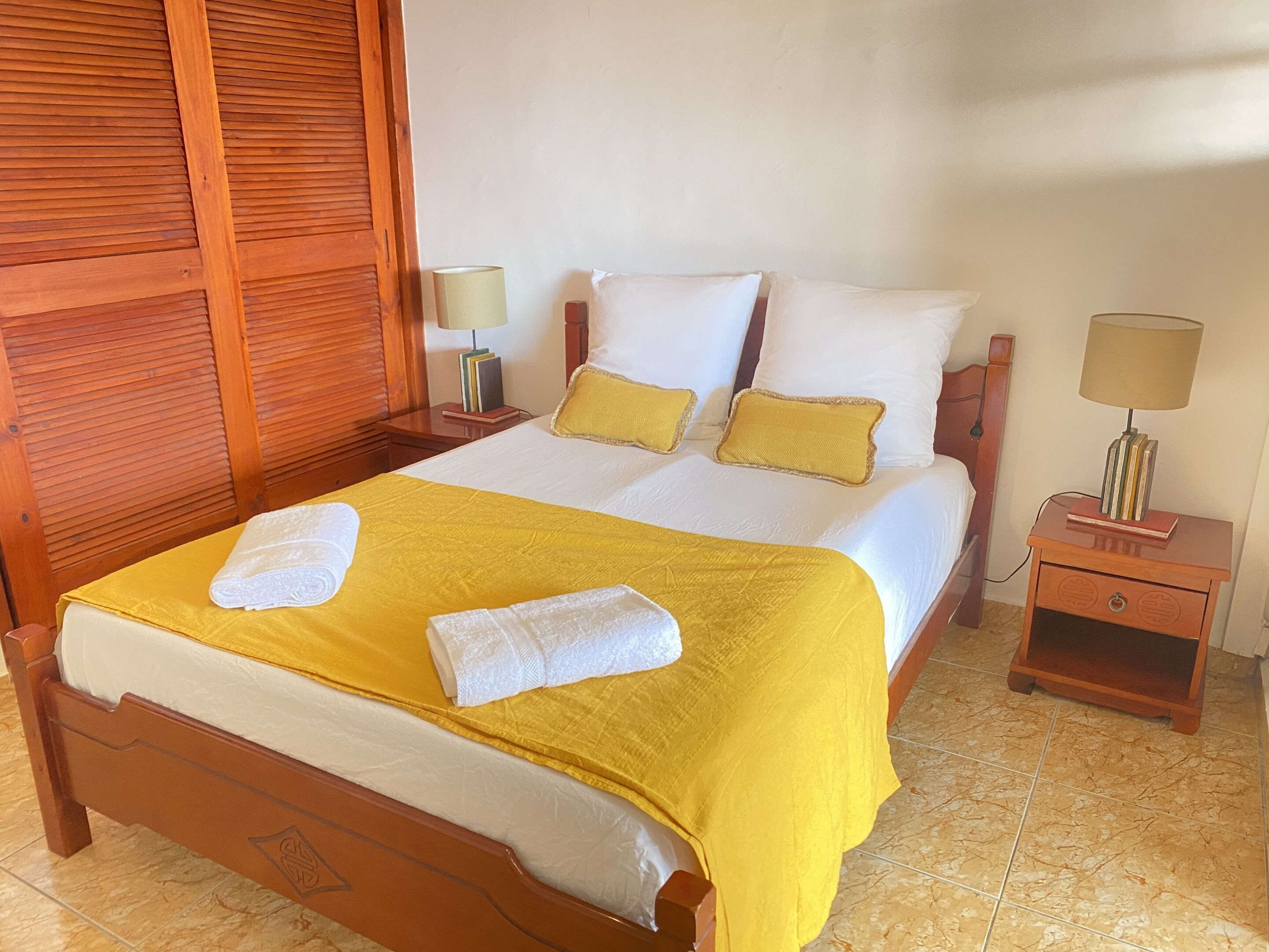 Cama bem arrumada com manta amarela, almofadas brancas e duas toalhas dobradas por cima. Há duas mesas de cabeceira com luminárias e livros.