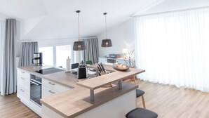 Uma cozinha moderna com ilha central, bancadas de madeira e banquetas. Há um forno embutido e uma cafeteira no balcão.
