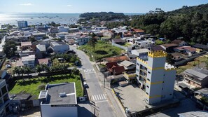 Vista aérea de uma cidade costeira com mistura de edifícios residenciais e comerciais, uma torre imponente com detalhes amarelos e uma visão clara do mar com barcos.