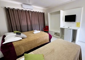 Quarto de hotel com cama, escrivaninha, cadeira, televisão e mini-geladeira.