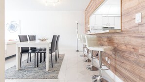 Uma área de refeições moderna com uma mesa e cadeiras brancas, um banco de bar e um espelho grande na parede.