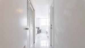 Corredor claro e branco que leva a uma sala de estar com sofá e janela.