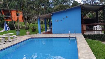 Uma piscina com deck ladrilhado, rodeada por vegetação e um edifício azul com varanda coberta.