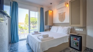 Quarto de hotel com uma cama, um pequeno frigorífico, janela com cortinas e vista para o exterior verde.
