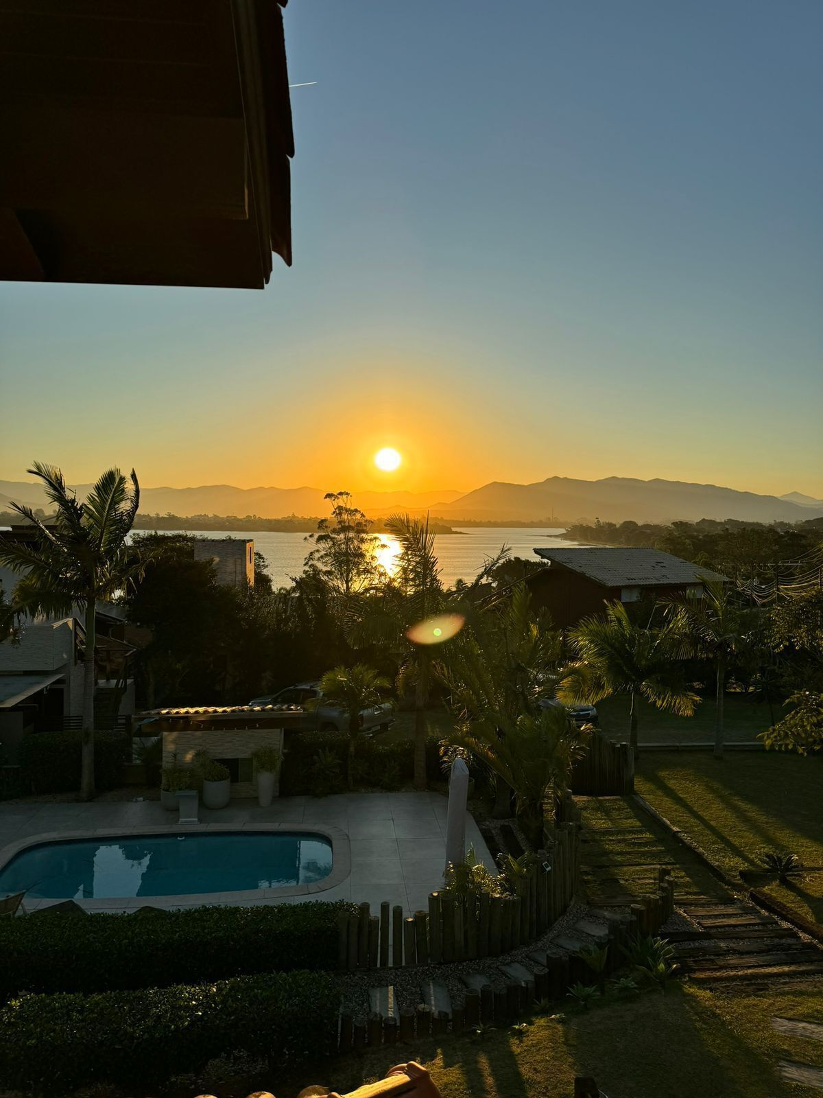 Área residencial com piscina, cerca de madeira e vista para o pôr do sol sobre a água e montanhas ao longe.