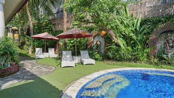 Piscina com borda azulejada, rodeada por grama sintética e uma parede de tijolos. Há duas espreguiçadeiras brancas e dois guarda-sóis.