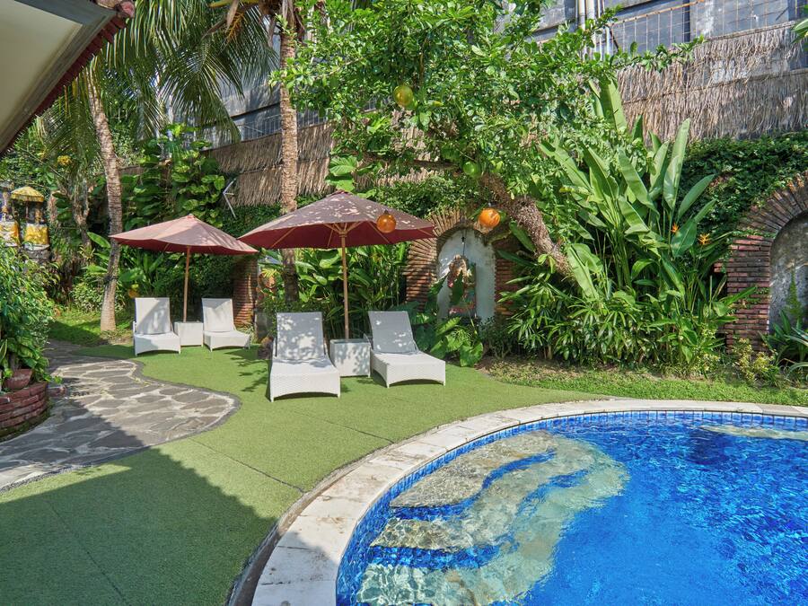 Piscina com borda azulejada, rodeada por grama sintética e uma parede de tijolos. Há duas espreguiçadeiras brancas e dois guarda-sóis.