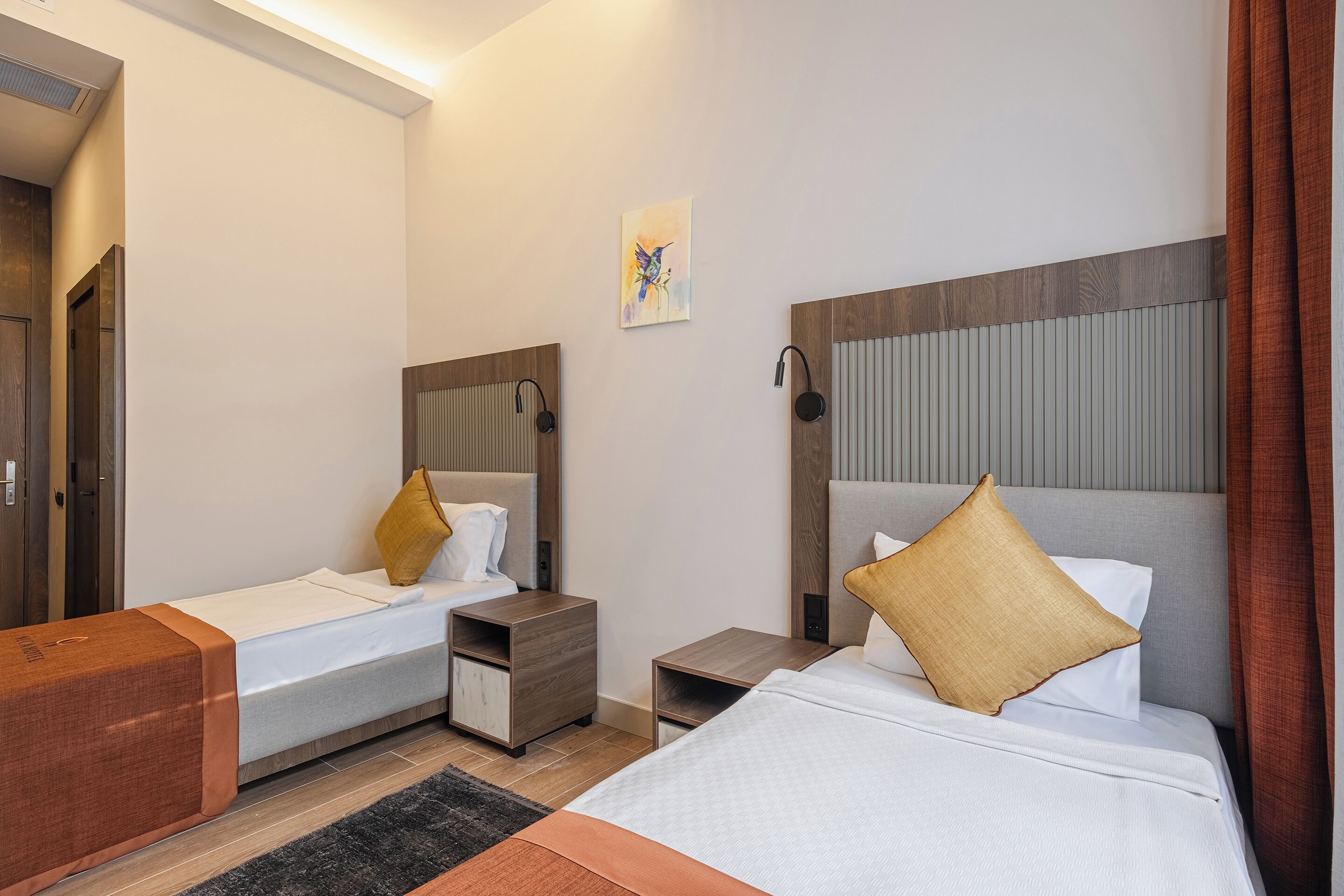 Quarto de hotel com duas camas de solteiro, uma pequena mesa de cabeceira com abajur, uma cama grande com cabeceira e um quadro na parede.