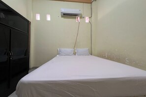 Uma cama de solteiro com roupa de cama branca num quarto com parede e ventoinha no teto brancos.