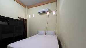 Uma cama de solteiro com roupa de cama branca, um ar condicionado fixado na parede e um guarda-roupa de cor escura.